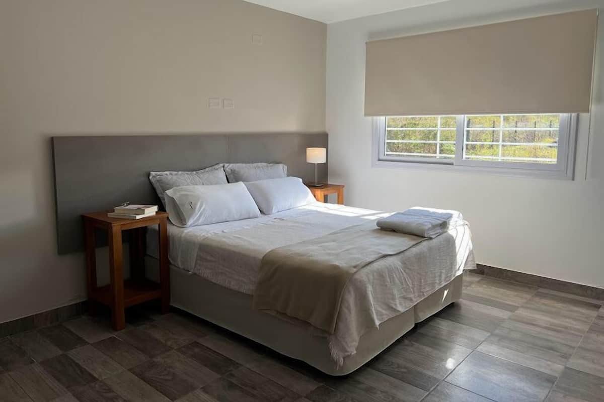 Quarto com cama, mesas de cabeceira, janela com persianas e vista para o verde lá fora.