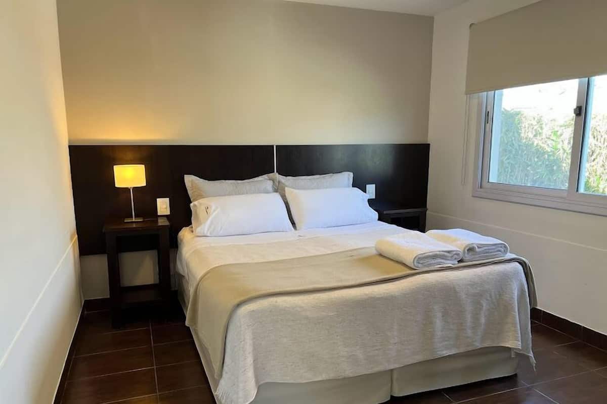 Cama bem arrumada com lençóis brancos, luminária de cabeceira e janela com vista para o verde.