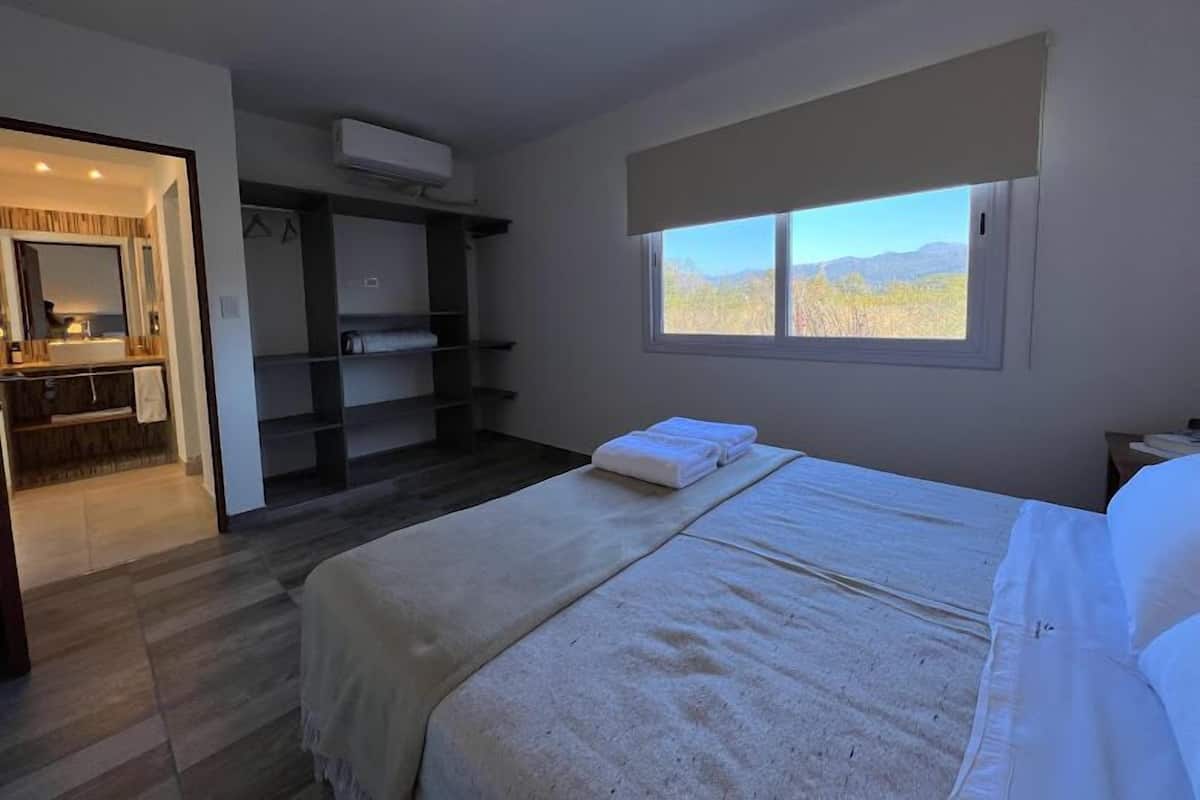Quarto com cama, janela com vista para as montanhas, armário embutido e banheiro com espelho e pia.