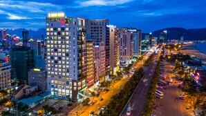 Um hotel alto com vários andares, uma placa grande no topo, e uma rua movimentada com carros e um ônibus em primeiro plano.