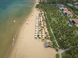 Vista aérea de um resort na praia, com uma longa fileira de guarda-sóis e espreguiçadeiras, uma piscina e uma série de edifícios do resort.