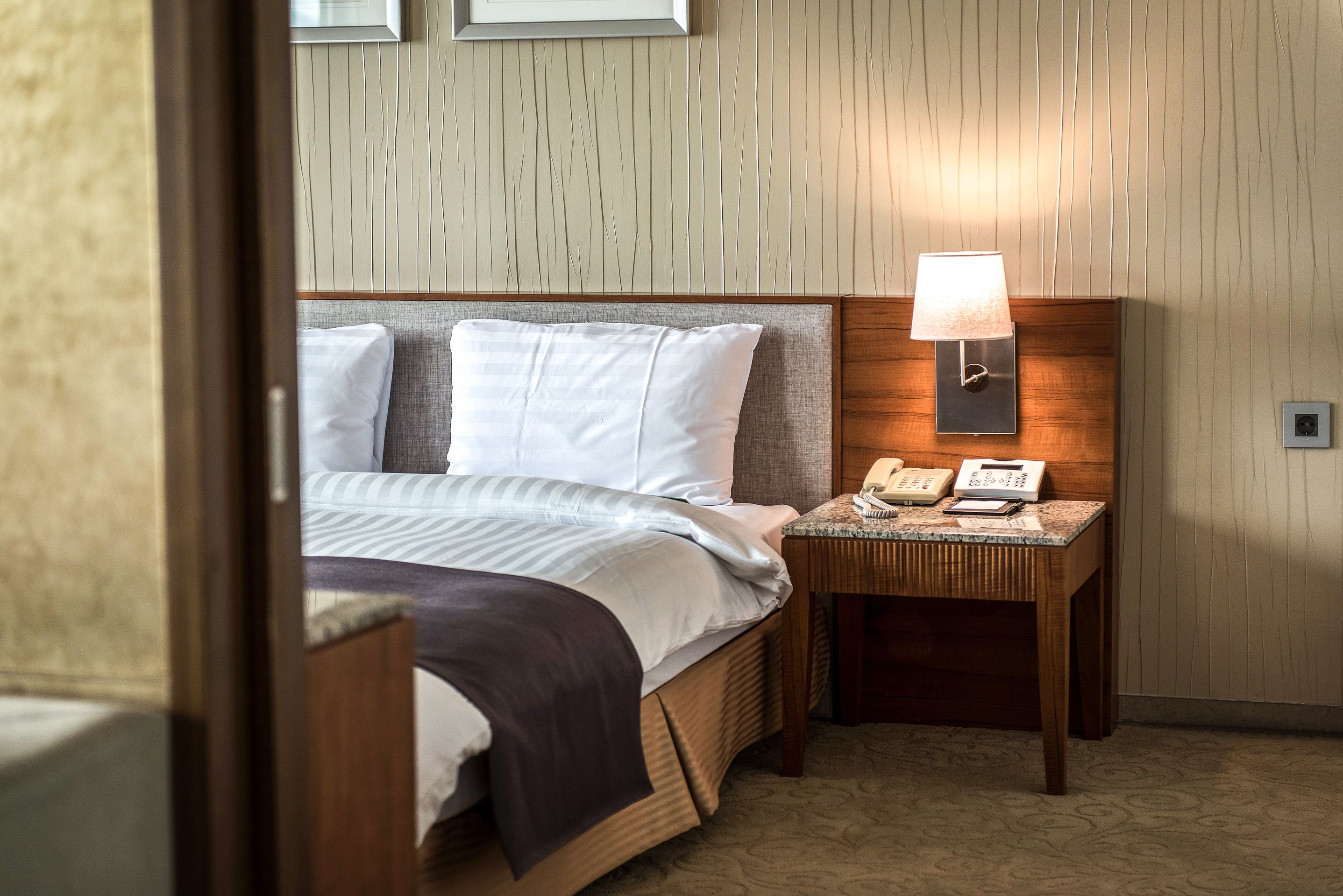 Quarto de hotel com cama, mesa de cabeceira com abajur e telefone.