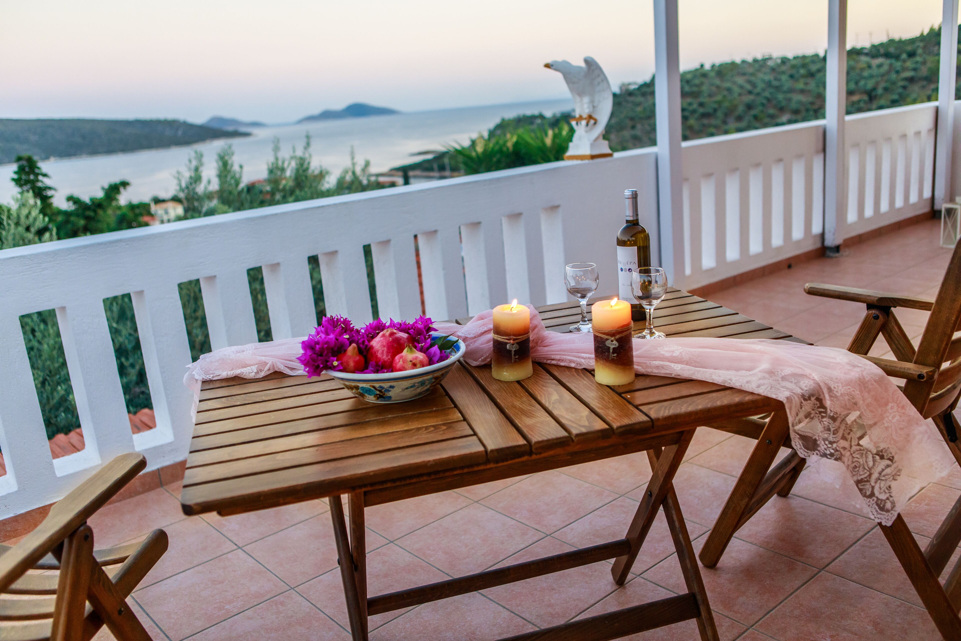 Uma mesa de madeira com uma tigela de frutas, velas e uma garrafa numa varanda com vista para a água.