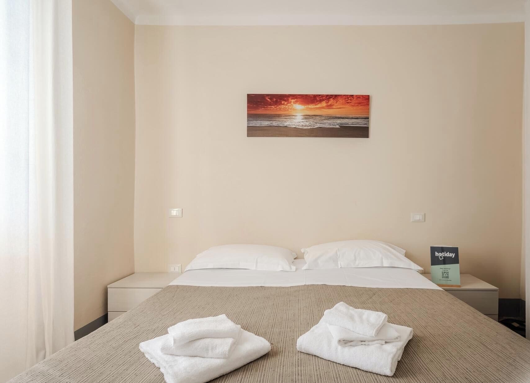 Quarto de hotel com cama, pintura do pôr do sol, placa indicando 'feriado' e toalhas na cama.