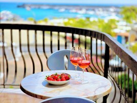 Uma mesa com uma tigela de morangos e dois copos de bebida vermelha, numa varanda com vista para o mar e edifícios.