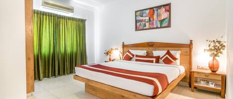 Quarto com cama de madeira, cortinas verdes, ventoinha no teto, ar condicionado fixado na parede, mesa de cabeceira com um vaso e um quadro na parede.