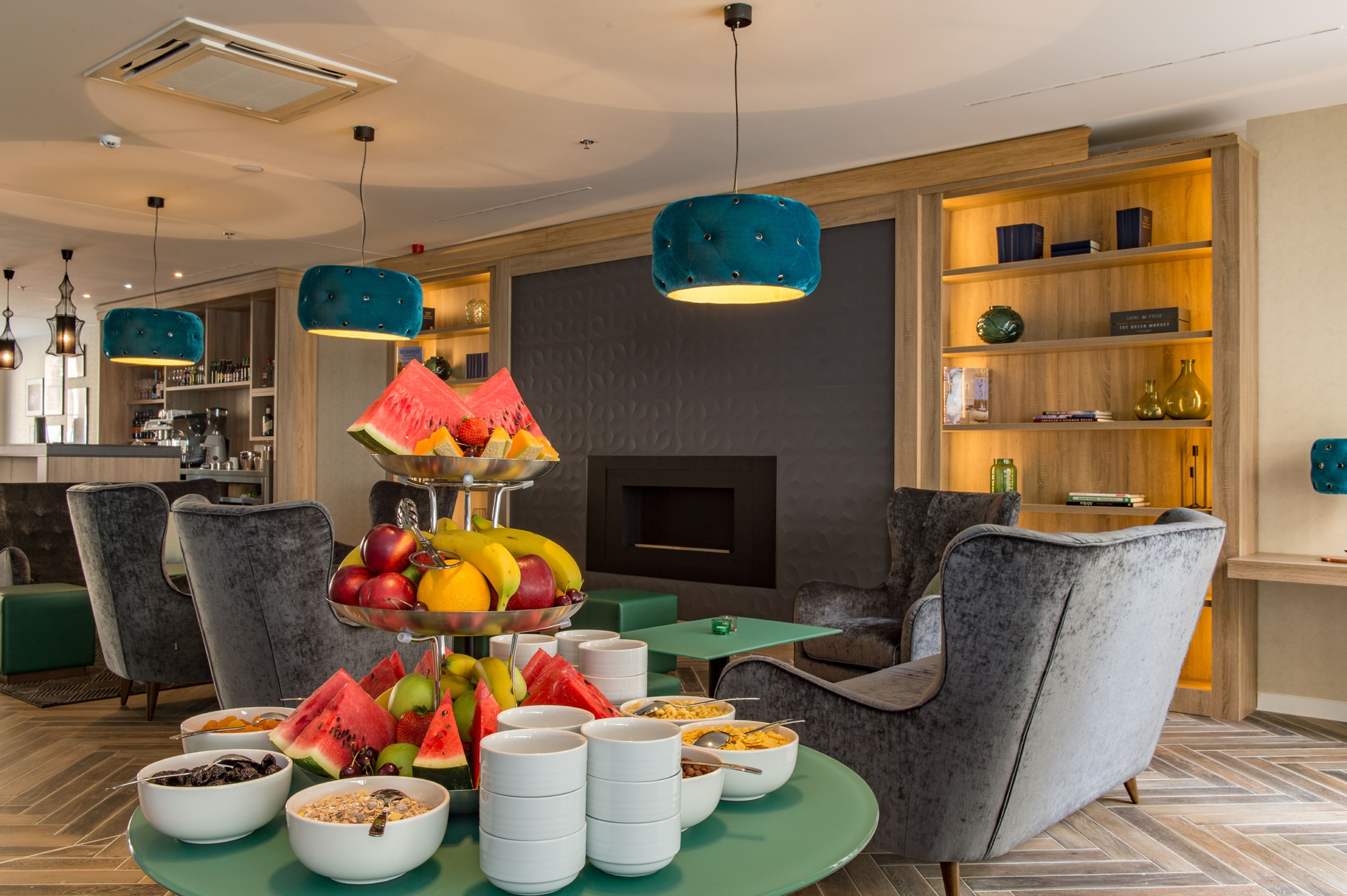 Uma área de refeições com buffet de frutas e cereais, cadeiras estofadas cinzas e um bar ao fundo.