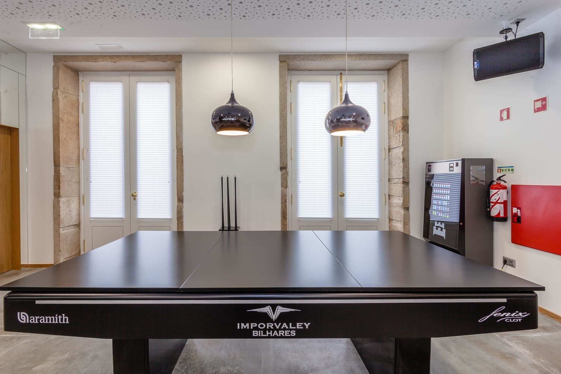 Mesa de bilhar com a marca "IMPORVALEY BILHARES", duas luminárias pendentes, um extintor de incêndio e uma máquina de vendas numa sala com paredes de pedra e grandes janelas.