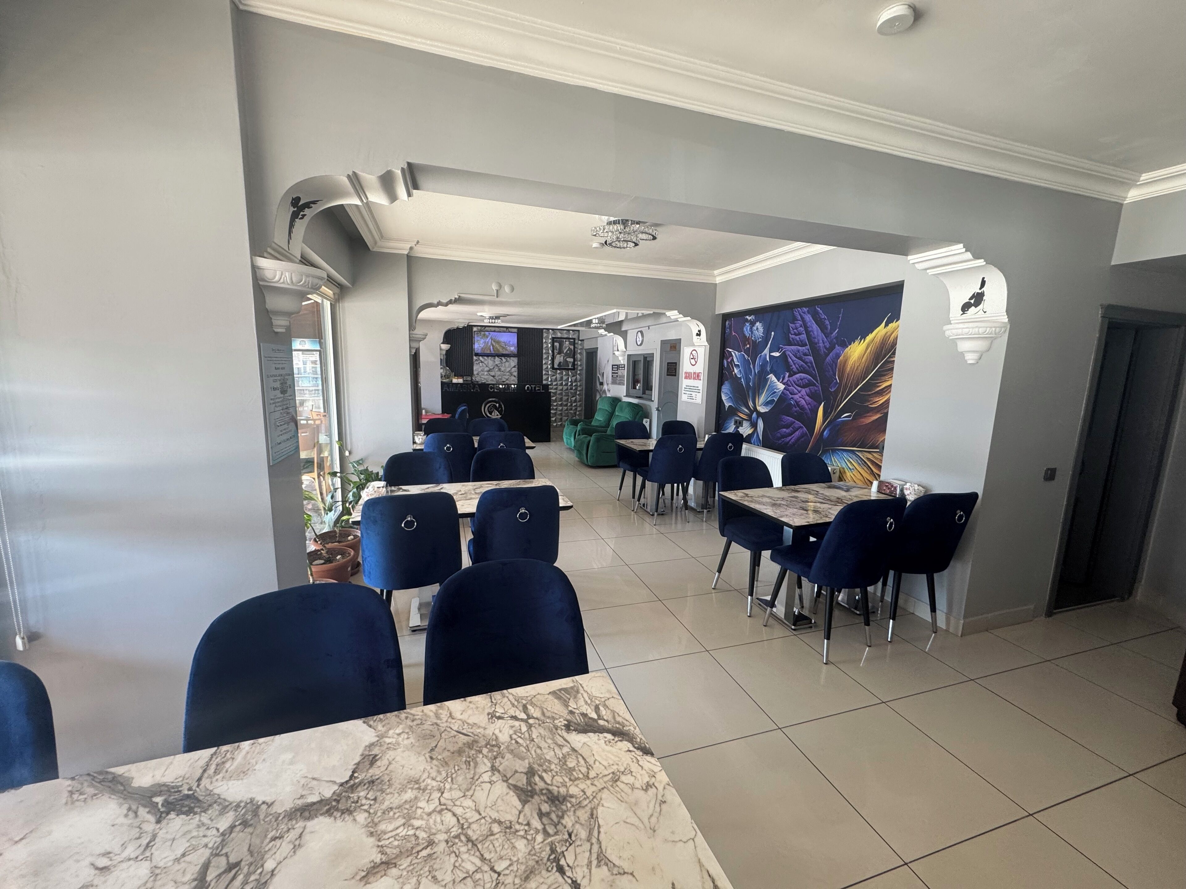 Uma área de refeições de hotel com mesas de granito e cadeiras azuis, um grande mural floral na parede e uma mesa de recepção ao fundo.