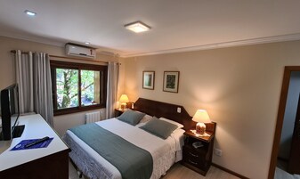 Quarto de hotel com cama, mesas de cabeceira, escrivaninha, televisão e janela com cortinas.
