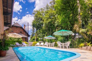 Piscina com espreguiçadeiras e guarda-sóis, cercada por vegetação exuberante e um edifício de tijolos.
