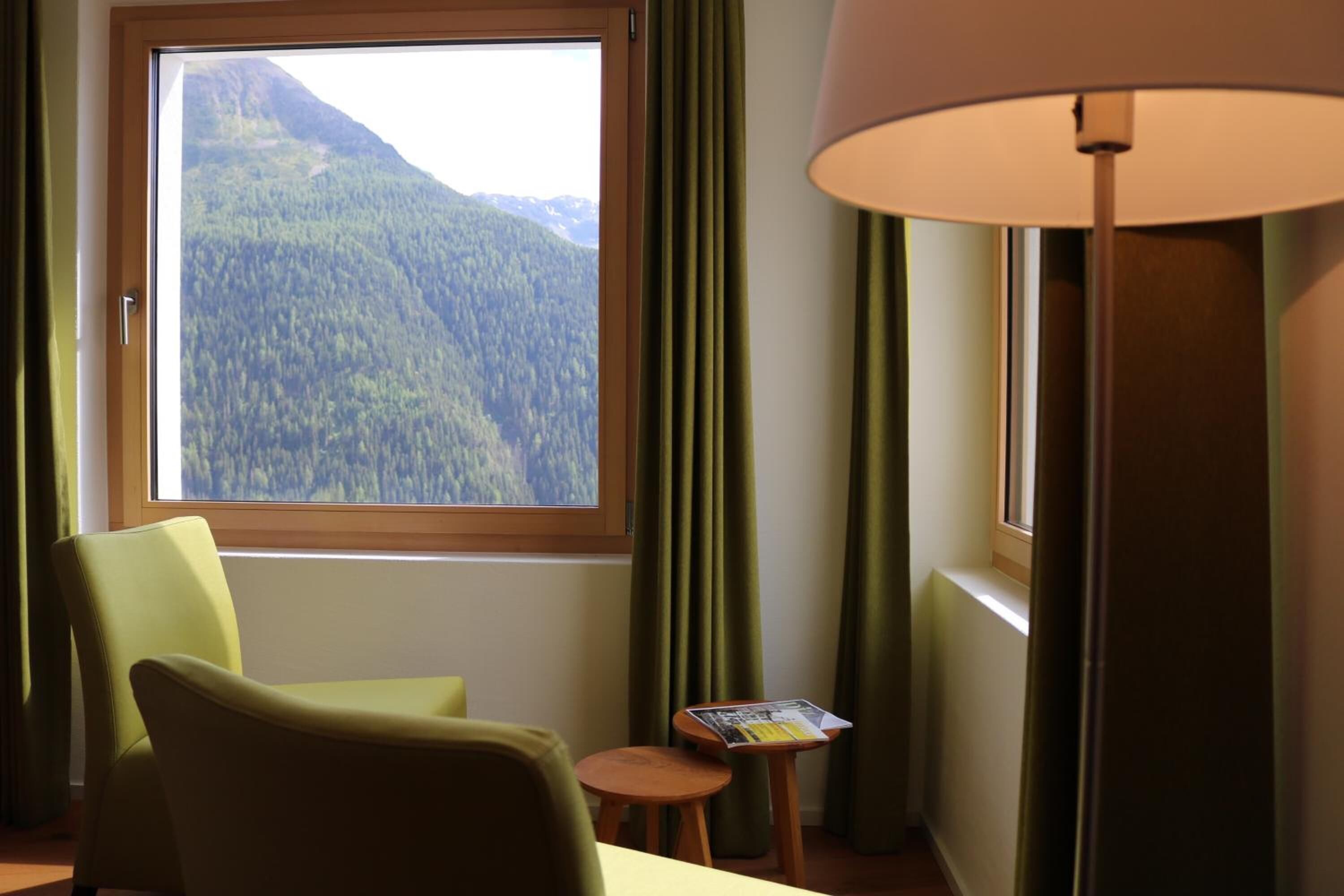 Quarto com uma janela ampla que oferece vista para as montanhas, uma cadeira verde, uma mesinha com uma revista e um abajur.
