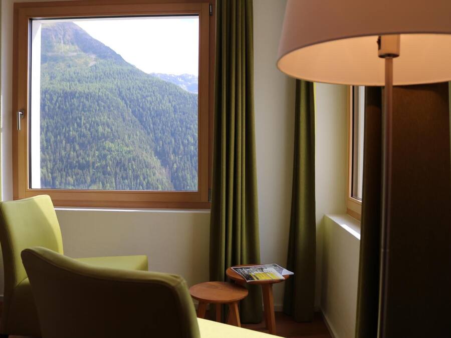 Quarto com uma janela ampla que oferece vista para as montanhas, uma cadeira verde, uma mesinha com uma revista e um abajur.