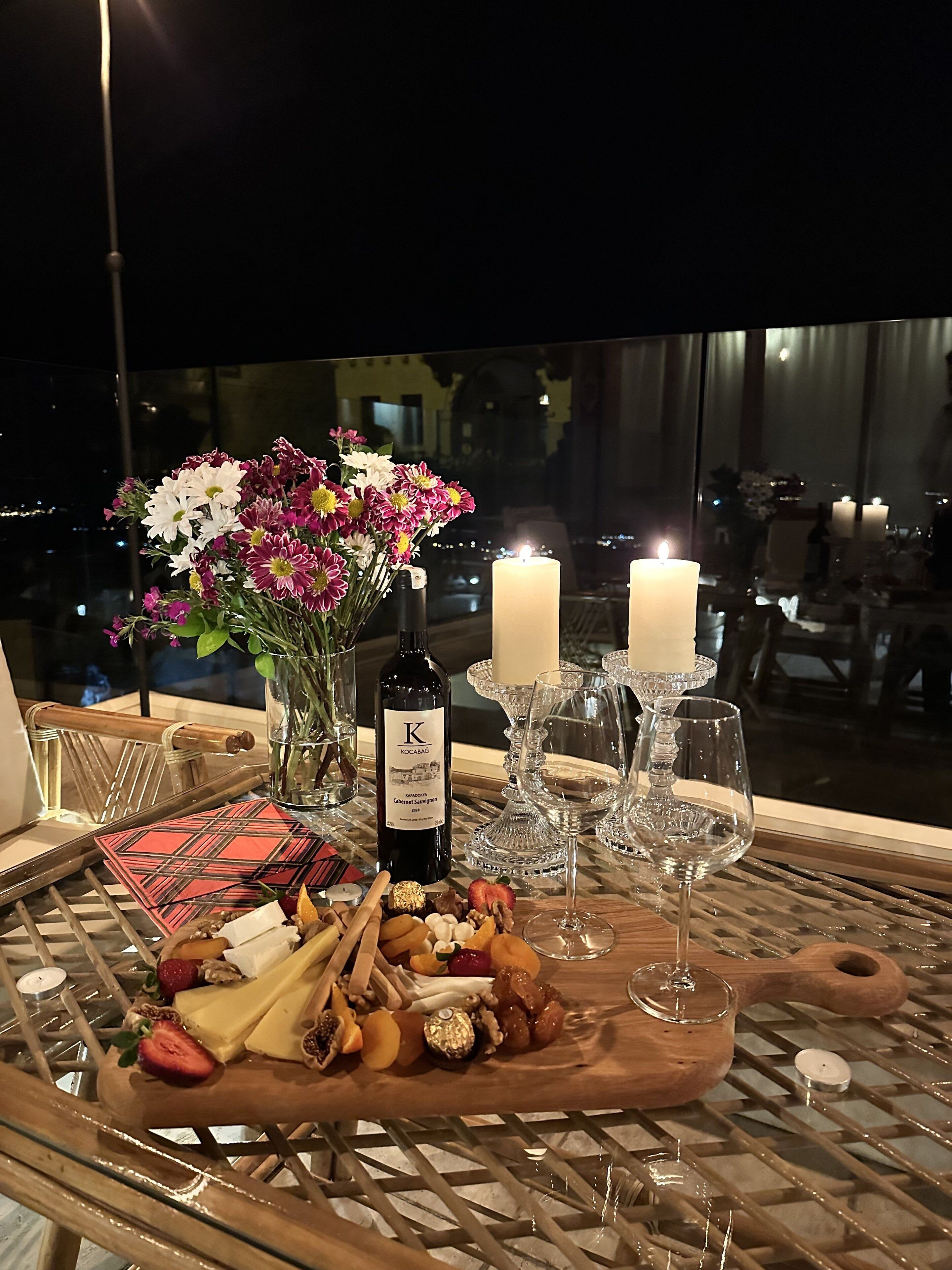 Mesa posta para refeição com garrafa de vinho, velas e variedade de frutas e queijos.