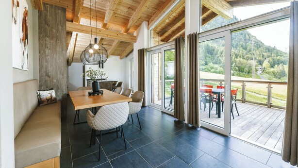 Uma área de refeições moderna com mesa e cadeiras de madeira, grandes janelas com cortinas e vista para um deck e montanhas.