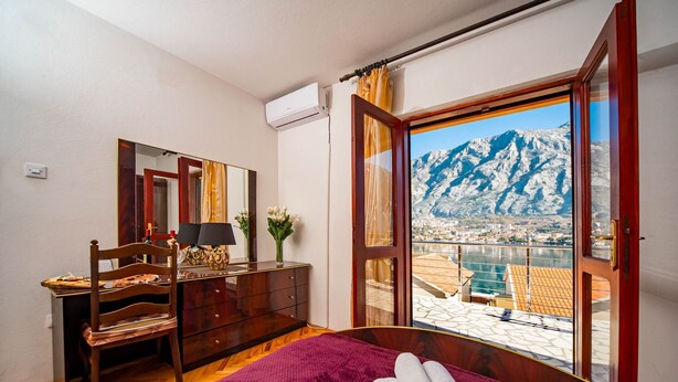 Quarto com cama, cômoda, cadeira, espelho e janela com vista para as montanhas.