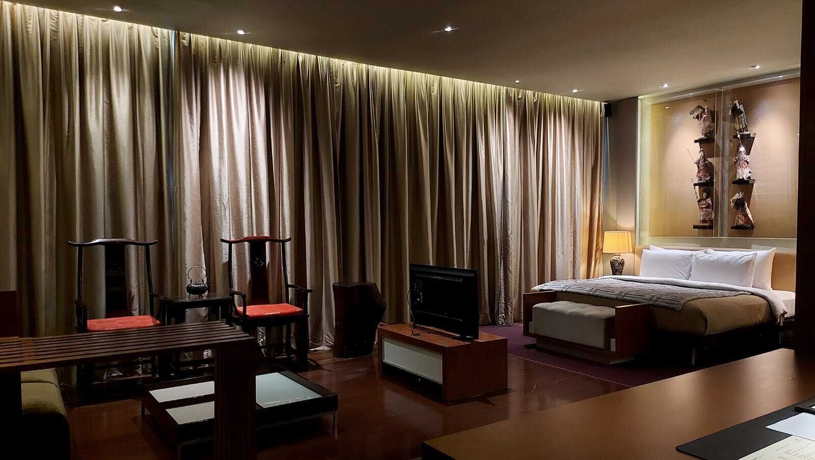 Quarto de hotel com uma cama grande, televisão, escrivaninha com cadeira e uma janela ampla com cortinas.