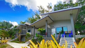 Um edifício moderno com entrada coberta, escadaria que leva à entrada principal e um jardim bem cuidado com plantas tropicais.