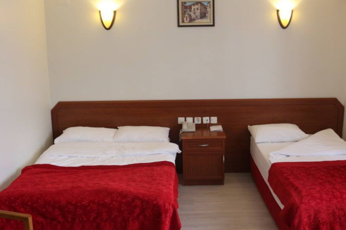Quarto com duas camas, cada uma com colcha vermelha e travesseiros brancos. Entre as camas, um criado-mudo de madeira com um telefone e uma mesinha.