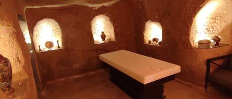 Um quarto com uma mesa central, três áreas de canto com iluminação e uma parede de pedra rústica.