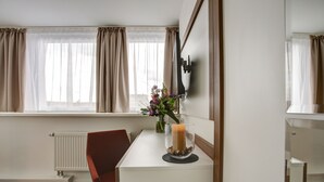 Quarto de hotel com uma secretária branca, uma cadeira vermelha, um vaso com flores e uma vela sobre a secretária.