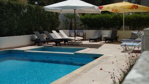 Piscina com espreguiçadeiras e guarda-sóis, cercada por vegetação e edifícios.