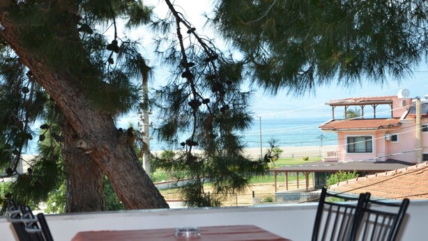 Mesa com toalha branca e duas cadeiras, com vista para a praia e um edifício com telhado vermelho.