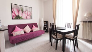 Uma sala de estar com um sofá roxo, uma mesa de jantar com cadeiras, uma estante de livros e uma janela com cortinas.