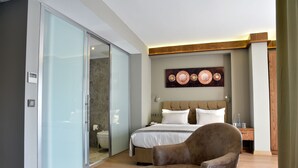 Quarto moderno com uma cama grande, uma porta de vidro deslizante para um banheiro e uma mesa de cabeceira de madeira com uma luminária.