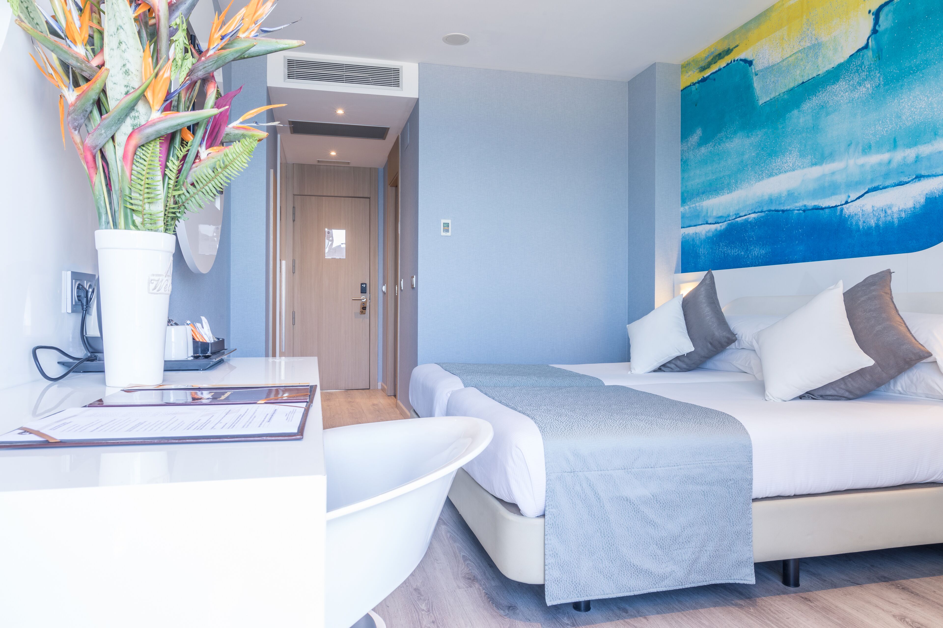 Quarto de hotel moderno com uma cama, uma mesa com uma planta e um quadro abstrato grande na parede.
