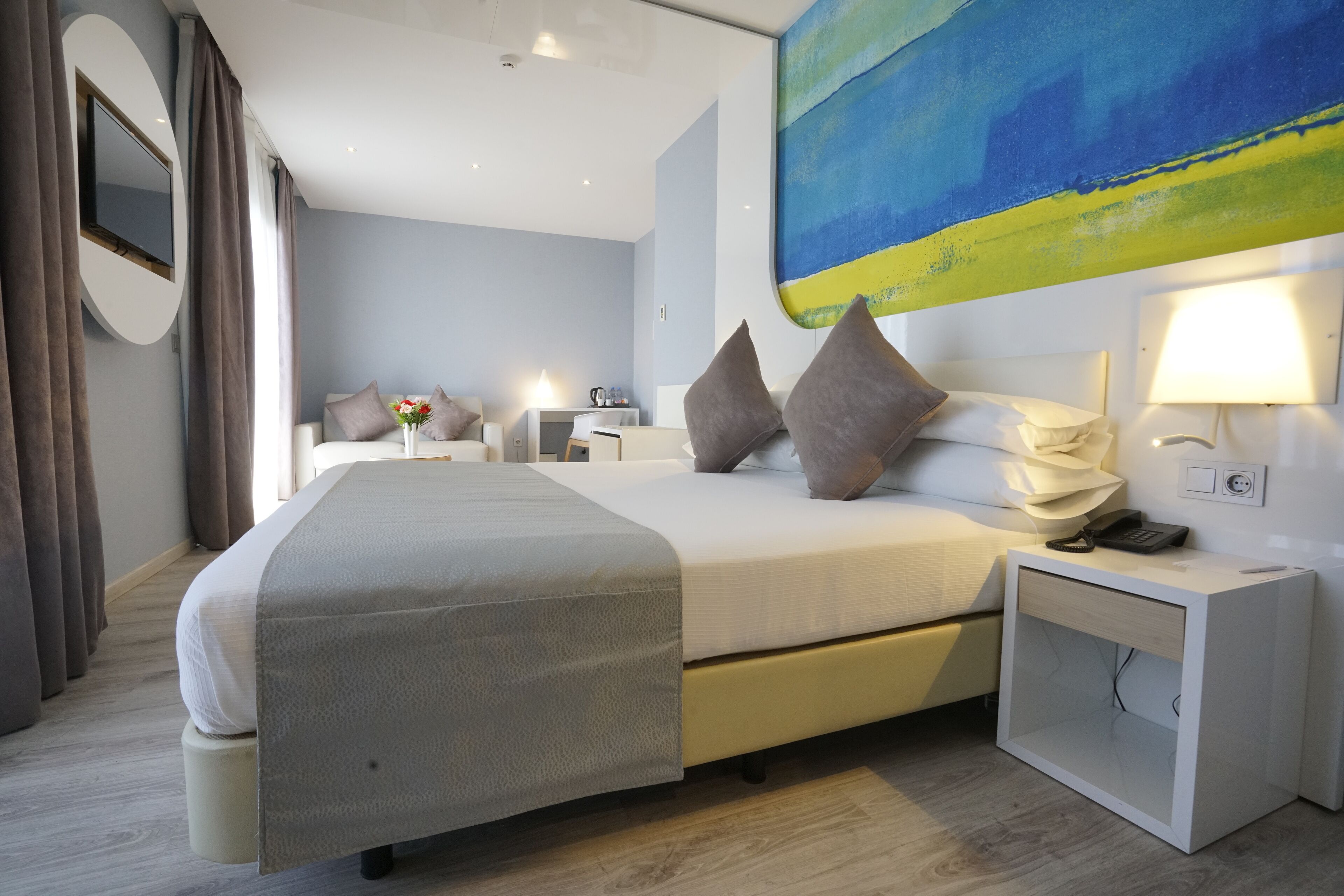 Quarto de hotel moderno com uma cama grande, mesas de cabeceira e um quadro na parede.