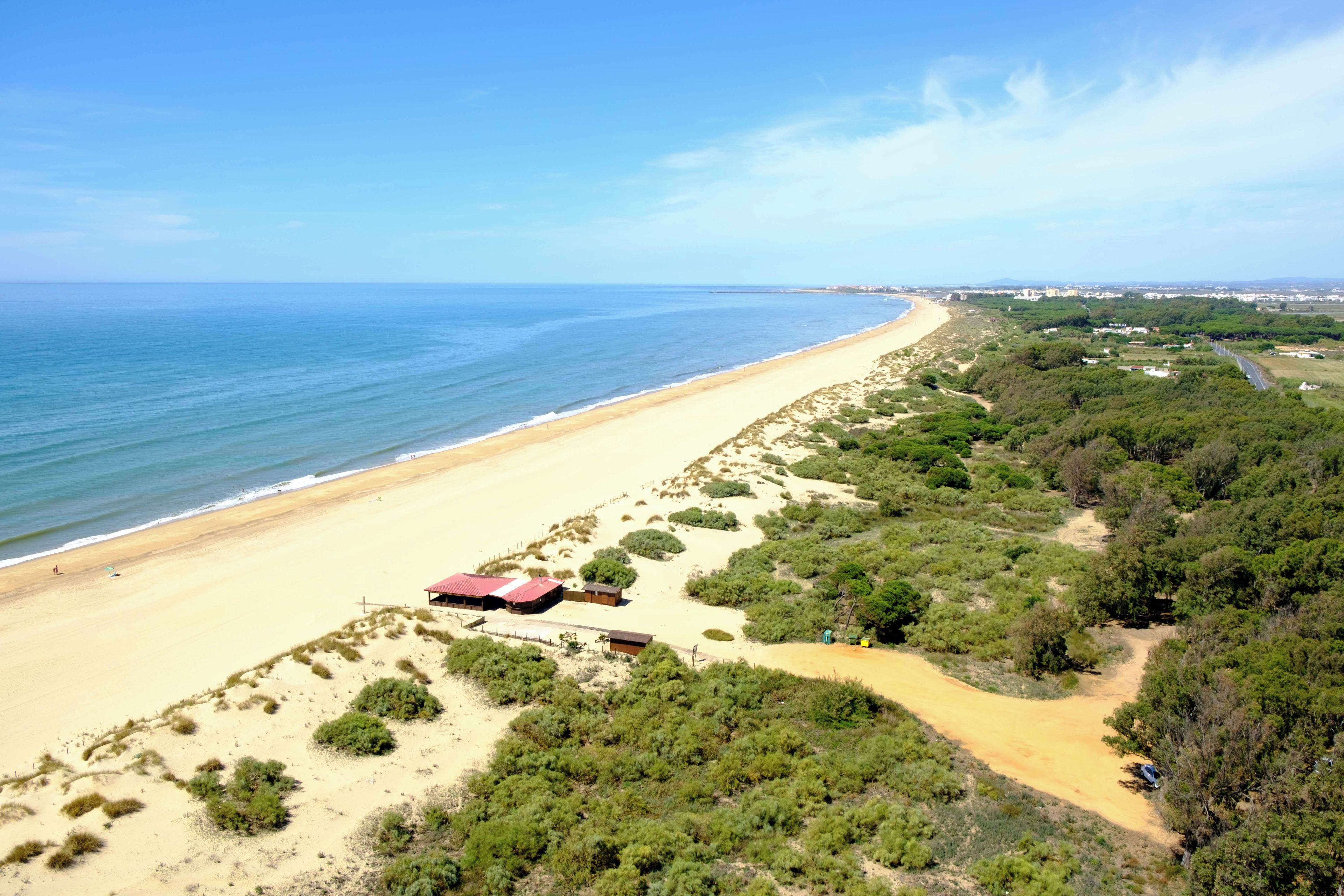 Paisagem costeira com uma praia arenosa, alguns edifícios e uma estrada serpenteando ao longo da costa.