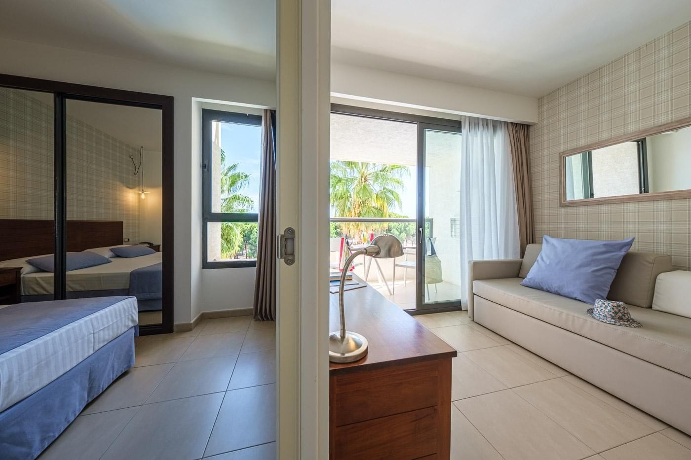 Quarto de hotel com uma cama, um sofá, uma mesa de apoio de madeira e uma porta de vidro deslizante para uma área externa com uma cadeira e uma mesa.
