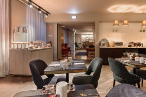Uma área de refeições de hotel com mesas preparadas para o café da manhã, um bar com petiscos e vista para a cozinha.
