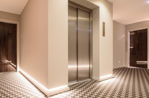 Um corredor com piso estampado, um elevador metálico e uma porta com o número 208.