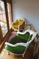 Quarto com duas cadeiras ornamentadas, uma amarela e outra verde, perto de uma grande janela com vista para um pátio.
