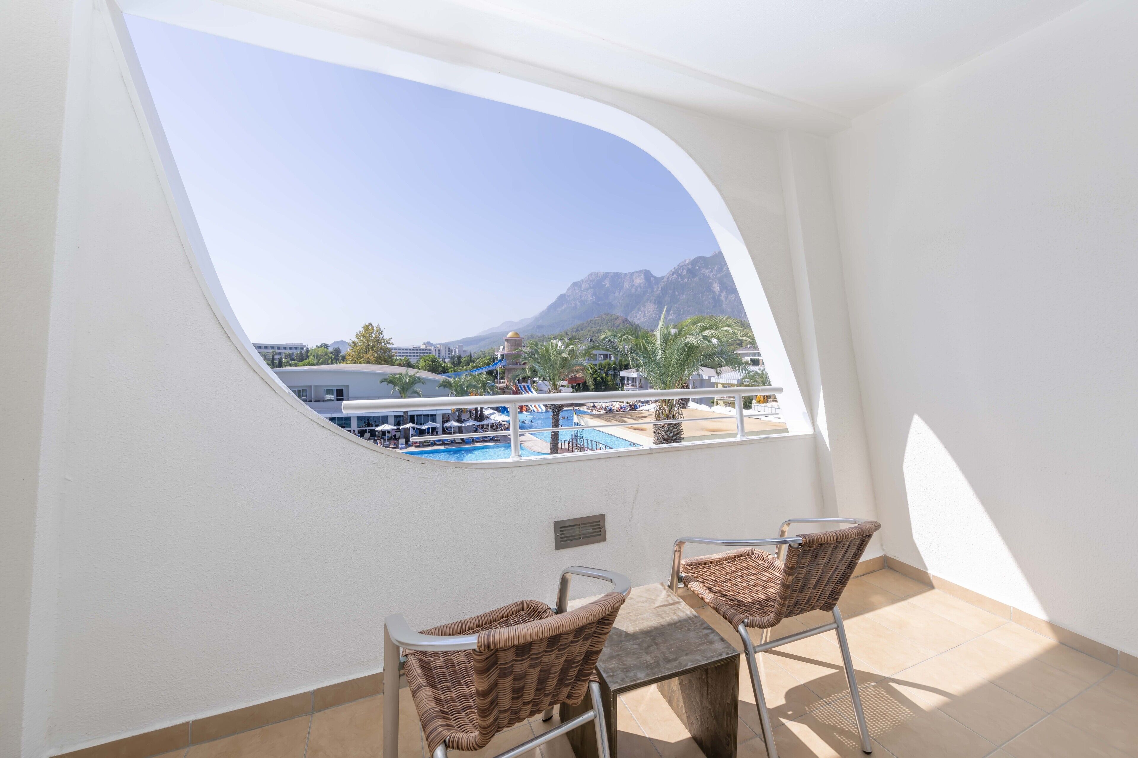Uma varanda com duas cadeiras de vime e uma mesa, com vista para uma área de piscina e uma montanha ao fundo.