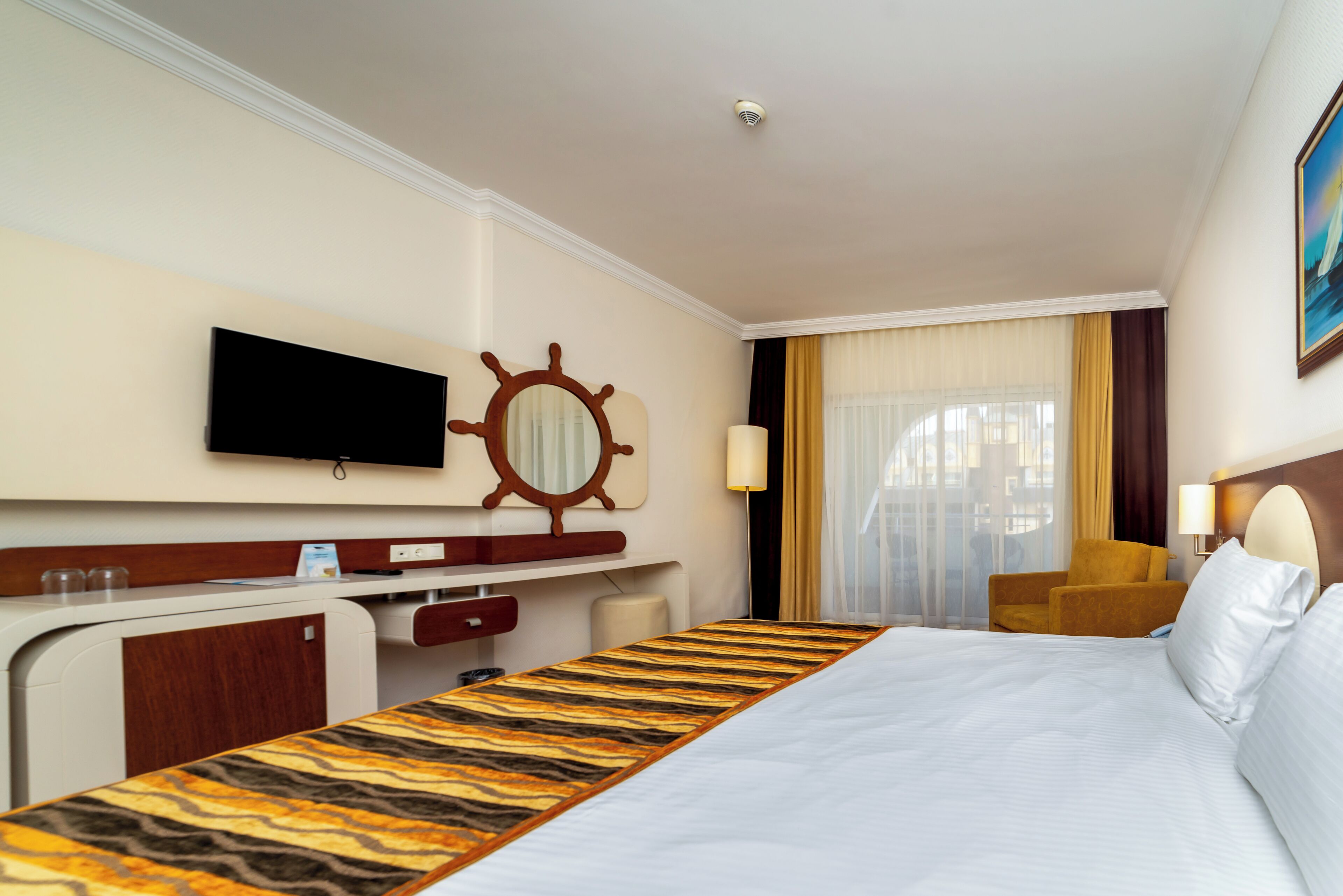 Quarto de hotel com uma cama grande, uma escrivaninha com televisão de tela plana, um espelho em formato de roda de navio, uma cadeira e uma janela com cortinas.
