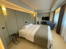 Quarto de hotel com cama grande, uma mesinha, televisão e área de estar com uma poltrona.