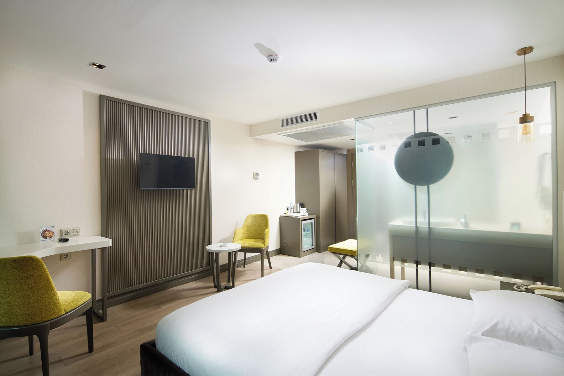 Quarto de hotel moderno com uma cama, escrivaninha, cadeira, televisão e divisória de vidro.
