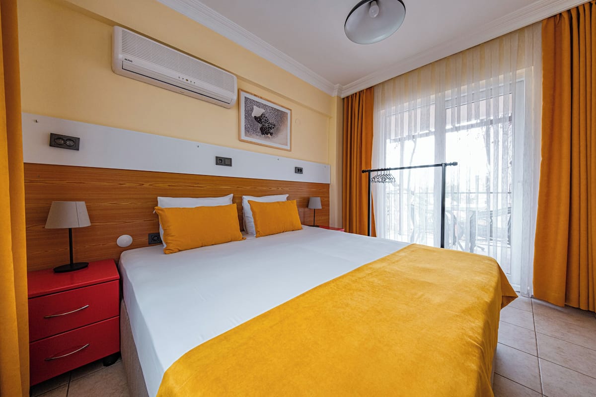Quarto de hotel com uma cama grande, uma mesa de cabeceira vermelha, ar-condicionado fixado na parede, um quadro emoldurado e uma janela com cortinas.