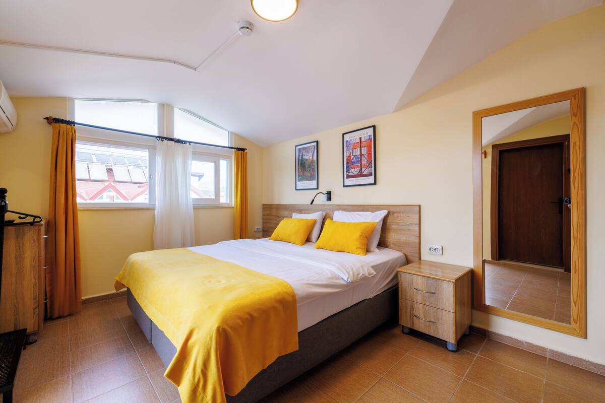 Quarto com cama, mesa de cabeceira, cômoda de madeira, janela com cortinas e porta.