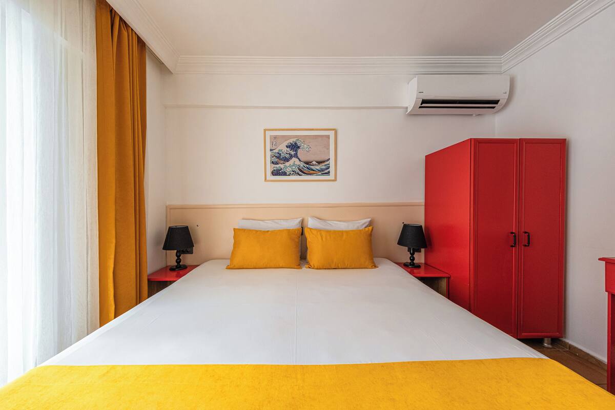 Quarto de hotel com uma cama grande, duas mesas de cabeceira com luminárias, um guarda-roupa vermelho, ar condicionado fixado na parede e uma obra de arte emoldurada sobre a cama.