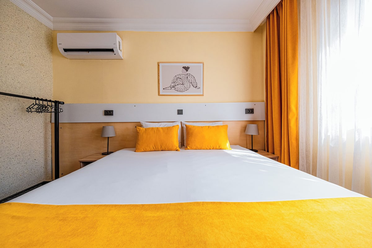 Quarto de hotel com uma cama grande, duas luminárias laterais, uma moldura na parede e um ar condicionado.