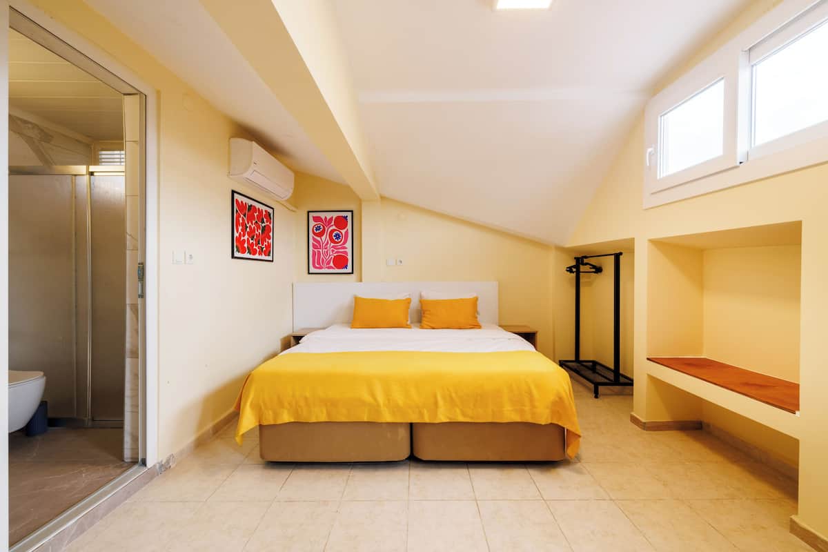 Quarto com cama, mesa de cabeceira, janela e porta de acesso a um banheiro.