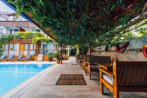Uma área à beira da piscina com espreguiçadeiras, uma mesa de madeira e um espaço coberto com vista para a piscina.