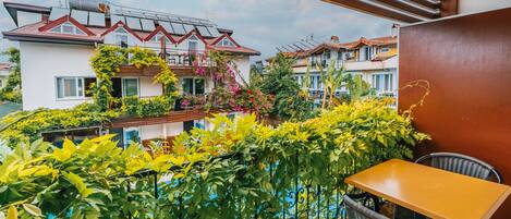 Varanda com mesa de madeira e cadeiras de vime, com vista para uma piscina e casas vizinhas.