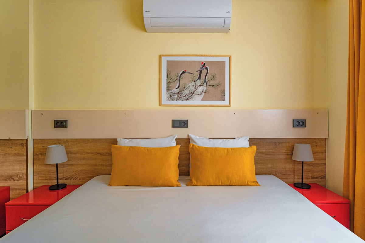 Quarto de hotel com uma cama grande, duas mesas de cabeceira, ar condicionado fixado na parede e um quadro emoldurado sobre a cama.