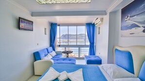Quarto de hotel moderno com uma janela ampla que oferece vista para o mar e montanhas, um sofá, uma cama com roupa de cama azul e uma pequena mesa.