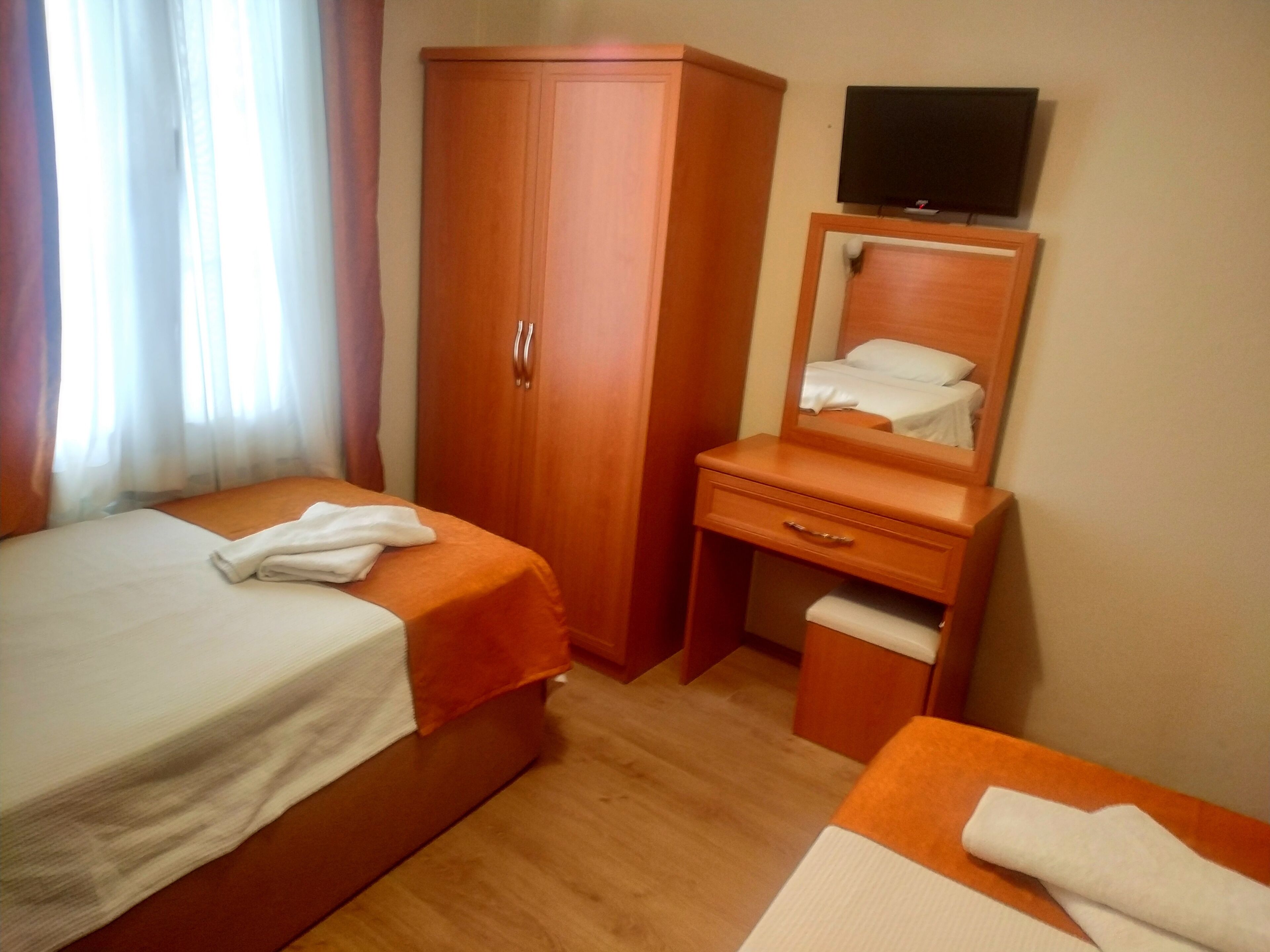 Quarto de hotel com duas camas, um guarda-roupa de madeira, uma secretária pequena com espelho e uma televisão na parede.